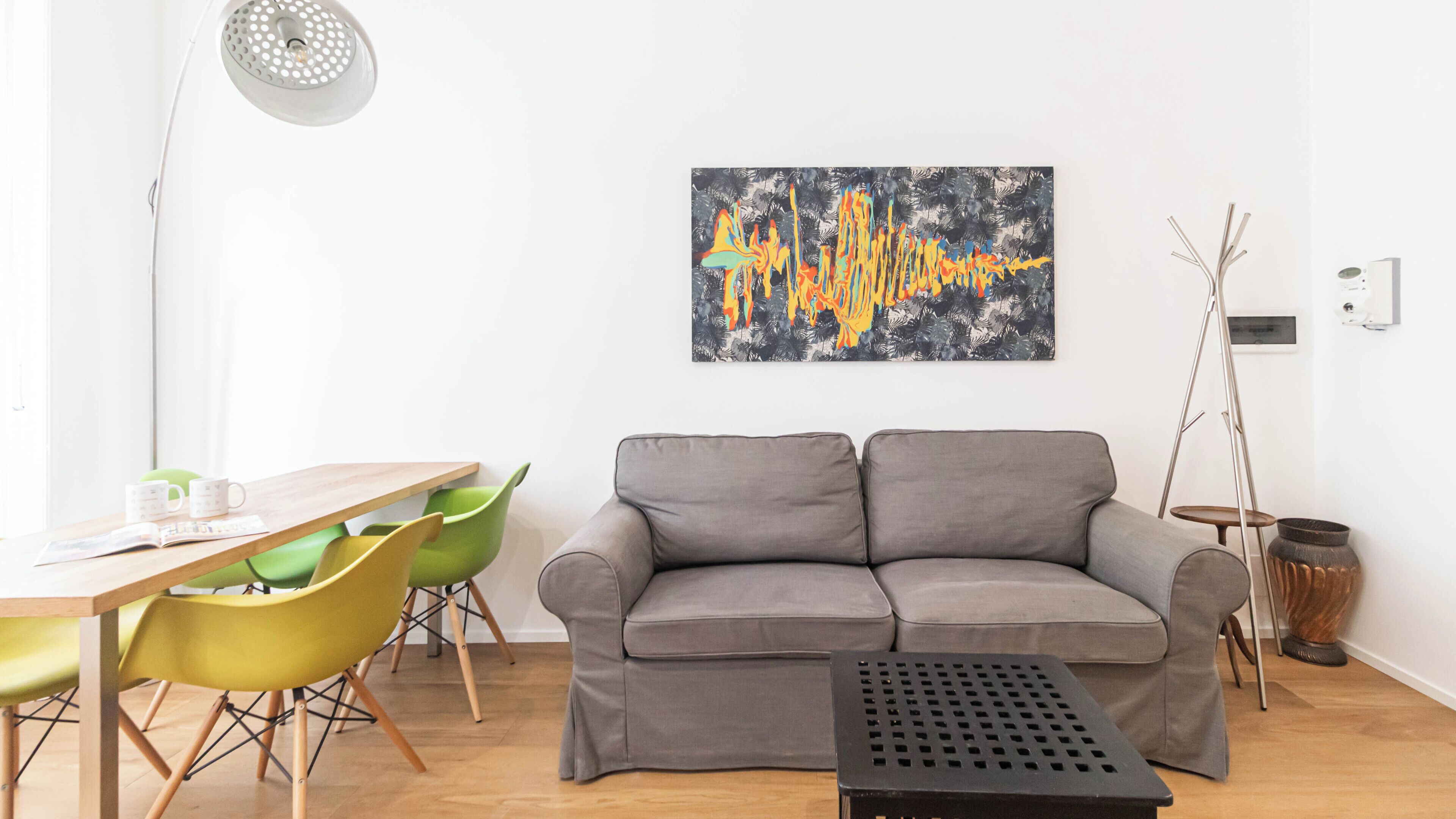 Uma sala de estar com um sofá cinzento, uma mesa de centro de madeira e uma poltrona verde. Há um quadro na parede e um abajur.