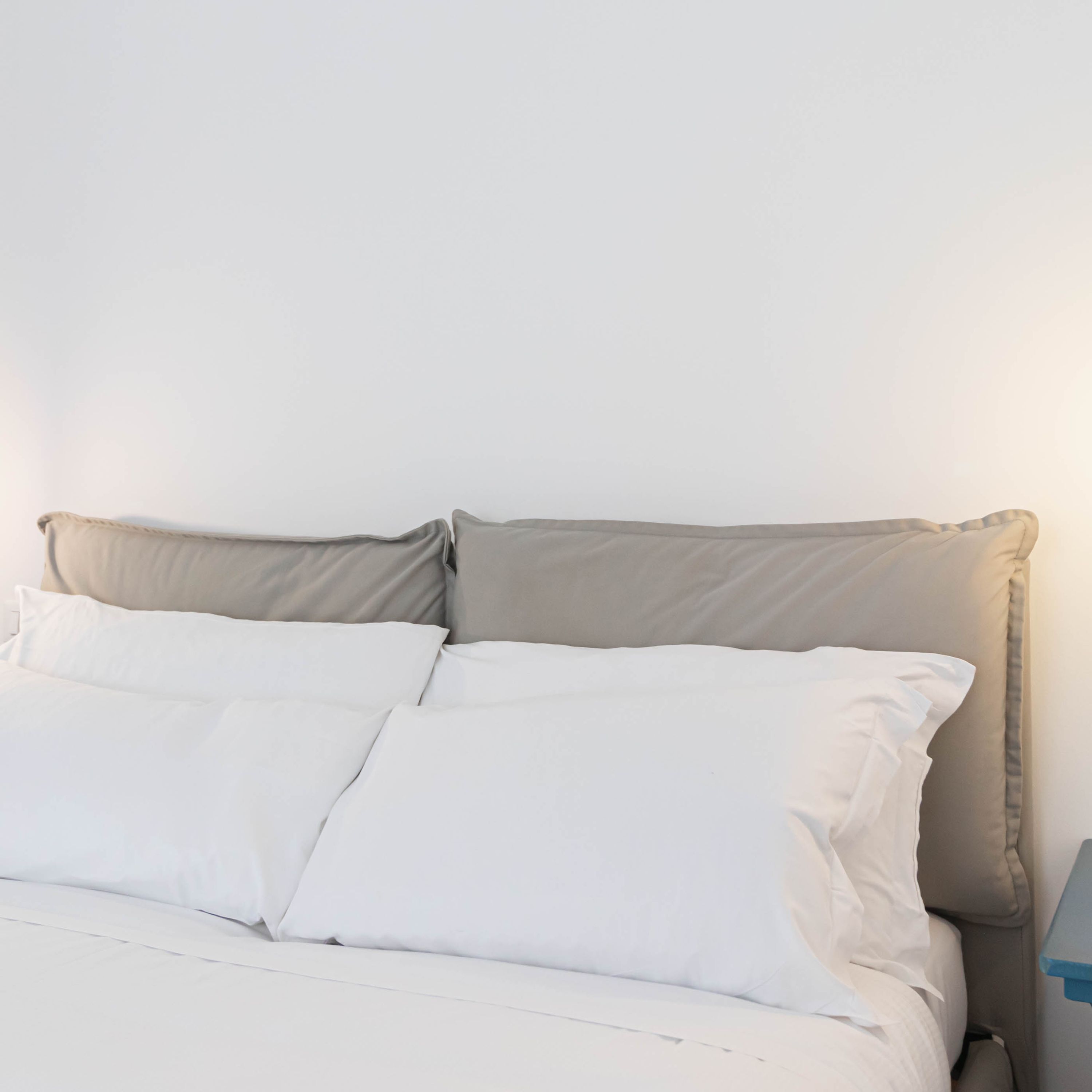 Uma cama com roupa de cama branca e bege, uma mesa de cabeceira azul e uma parede lisa.