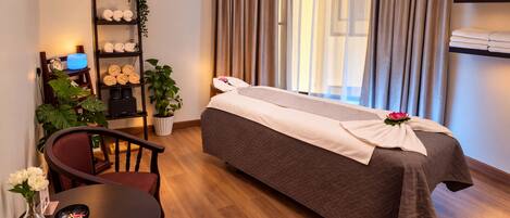 Quarto de hotel com cama, escrivaninha, cadeira, mesinha, planta e prateleira com toalhas e relógio.