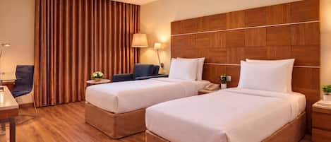 Quarto de hotel com duas camas, uma escrivaninha, uma cadeira, um abajur e uma cortina.
