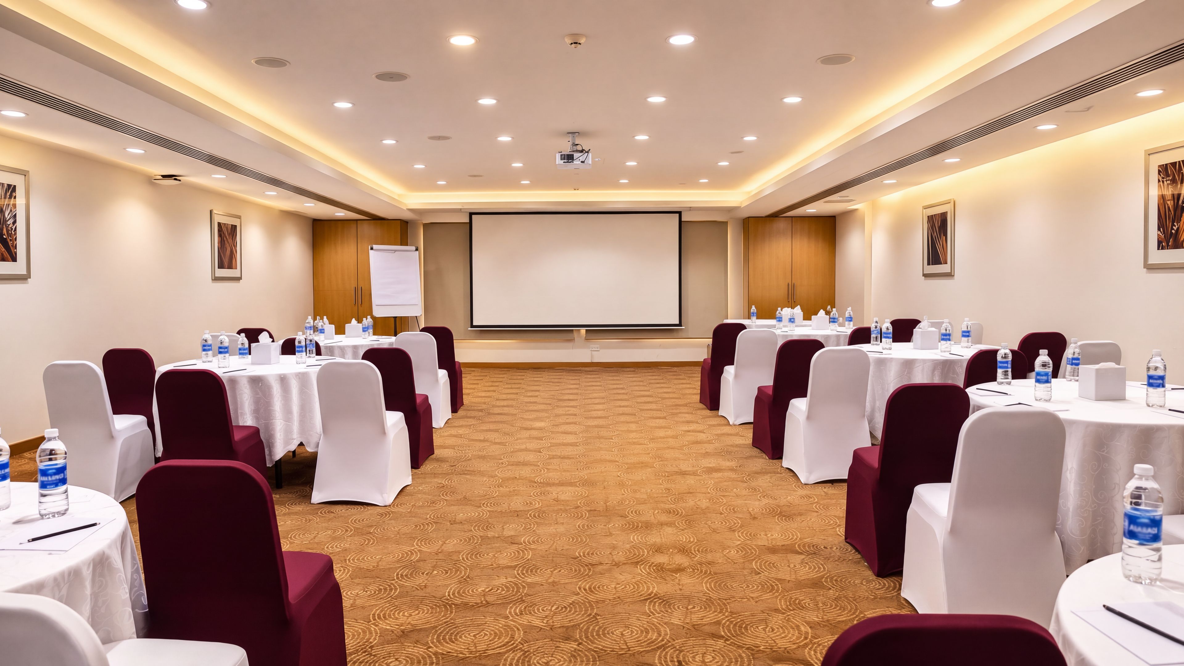 Sala de conferências com tela grande, mesas brancas com garrafas de água e cadeiras vermelhas.
