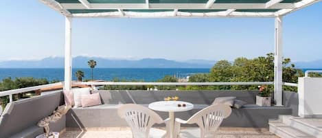 Área de estar coberta ao ar livre, com mesa e cadeiras, oferecendo uma vista panorâmica do mar e das montanhas.