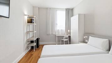 Quarto de hotel com duas camas, uma escrivaninha, uma cadeira, um guarda-roupa e uma janela com cortinas.