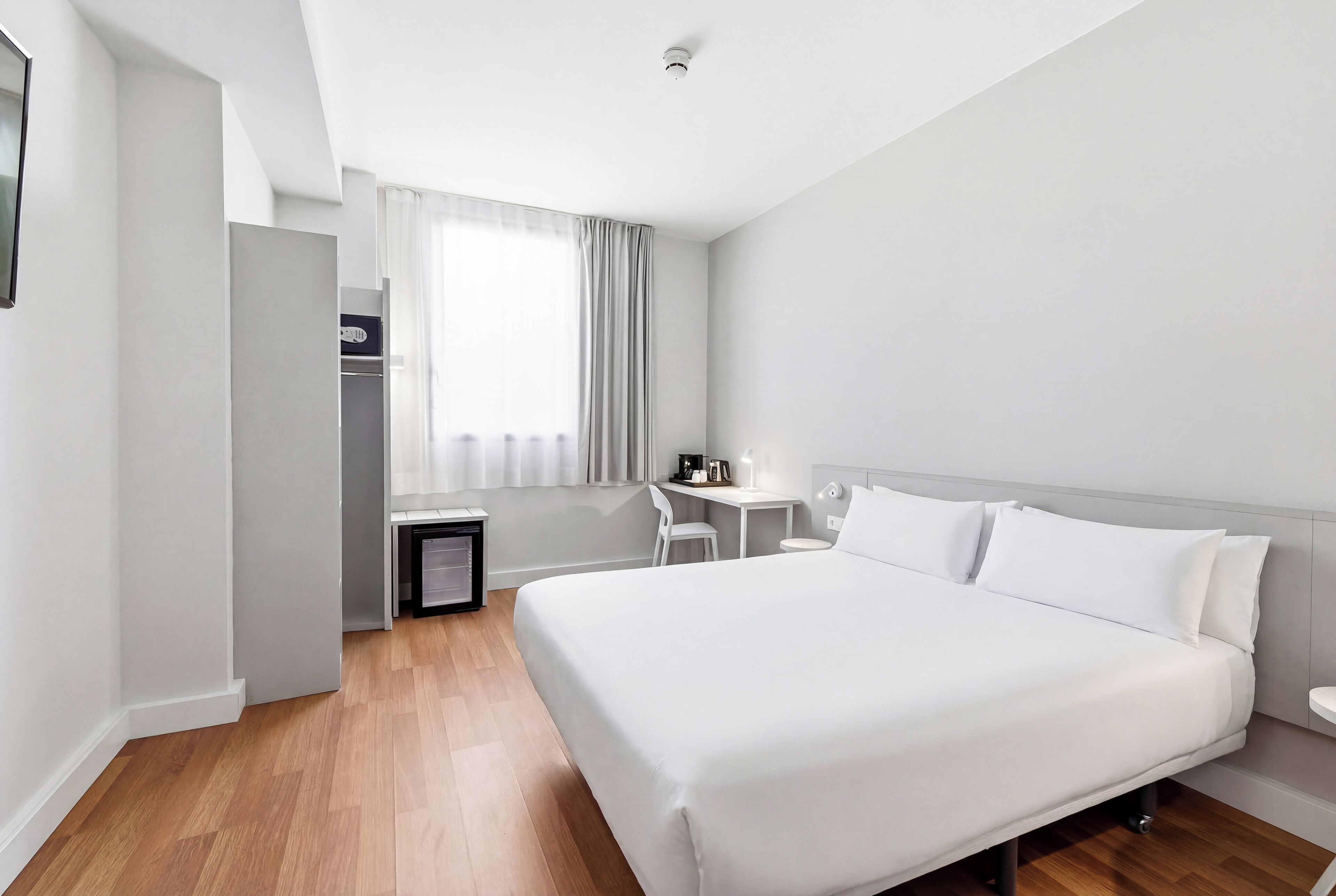 Quarto de hotel com cama, escrivaninha, cadeira, mesa pequena e guarda-roupa.