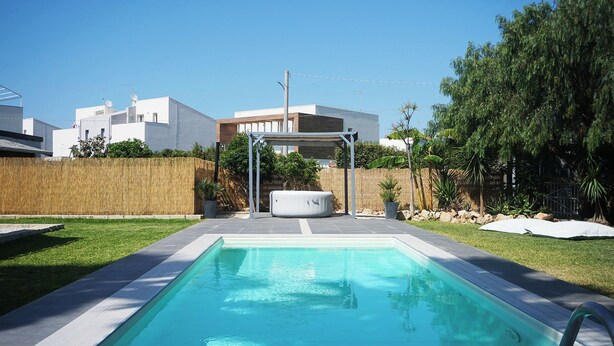 Uma piscina retangular com superfície de água azul cristalina, rodeada por um deck pavimentado. Há uma área de estar coberta com uma estrutura branca ao fundo.