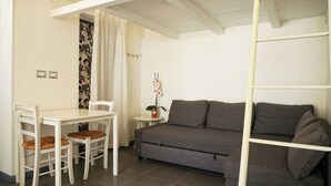 Quarto com uma mesa branca, duas cadeiras, um sofá cinza e uma pequena mesa lateral com uma planta em vaso.