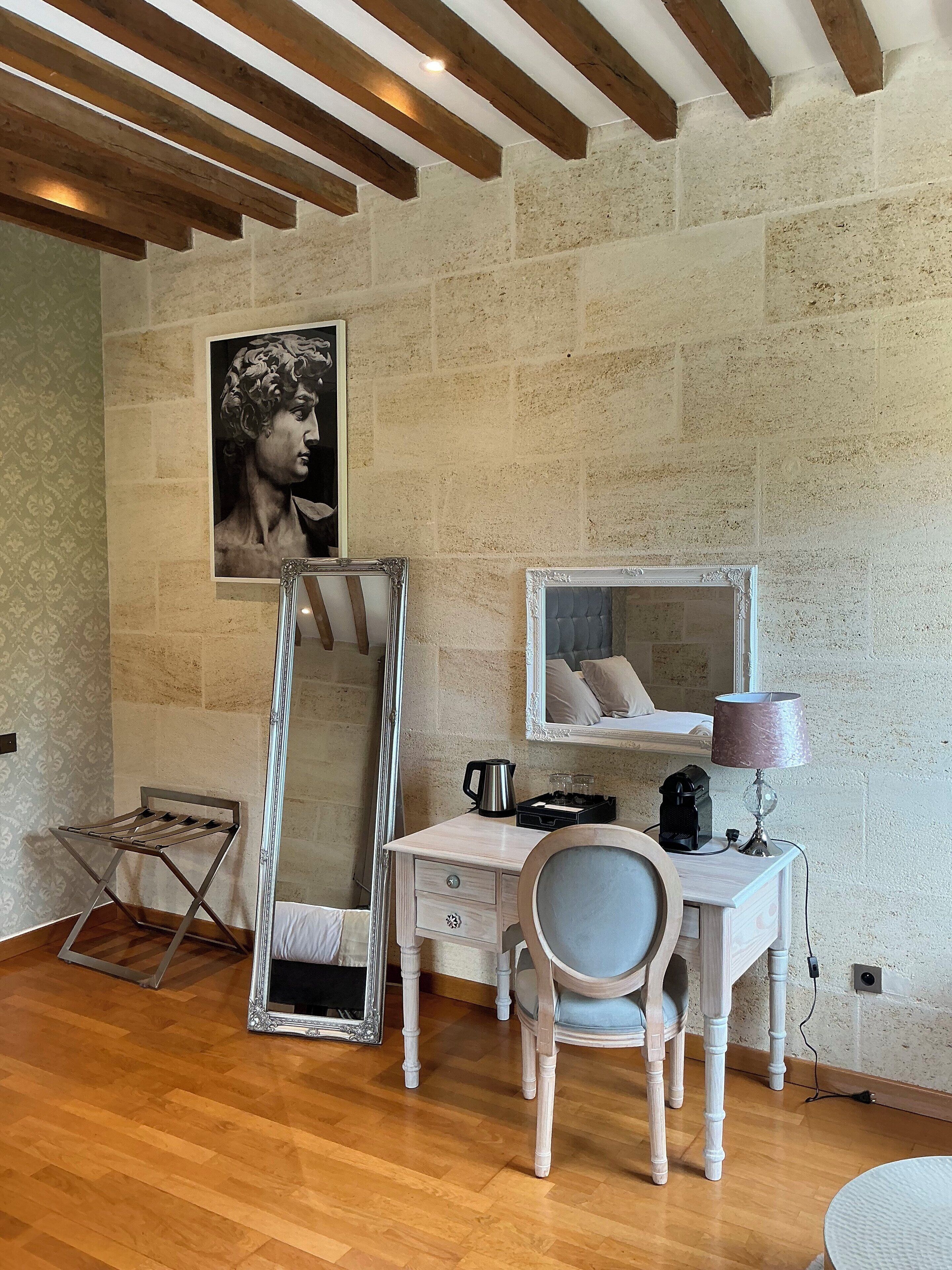 Quarto com teto de madeira, parede de pedra, chão de madeira, uma mesa branca com cadeira, espelho, abajur e quadro emoldurado.