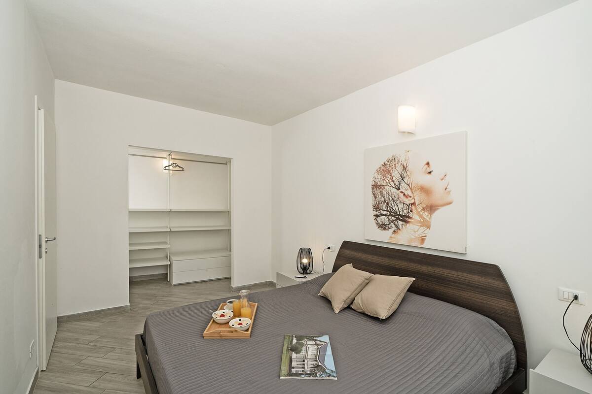 Quarto moderno com cama, mesa de cabeceira, luminária de parede, quadro e armário com cabides.