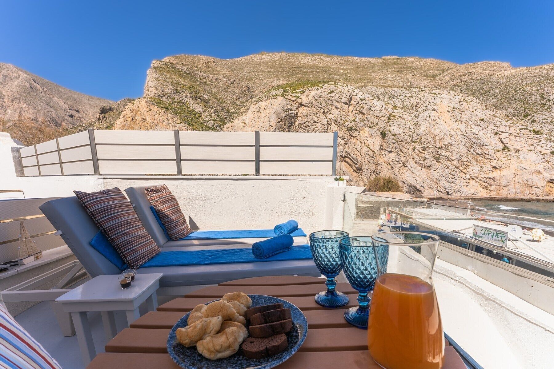 Um terraço com área de estar, uma mesa com comida e bebidas, e vista para o mar e montanhas.