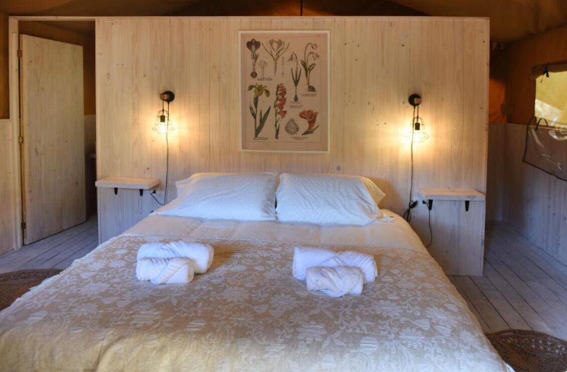 Quarto com teto de madeira, cama com lençóis brancos, duas luminárias de cabeceira e uma ilustração botânica na parede.