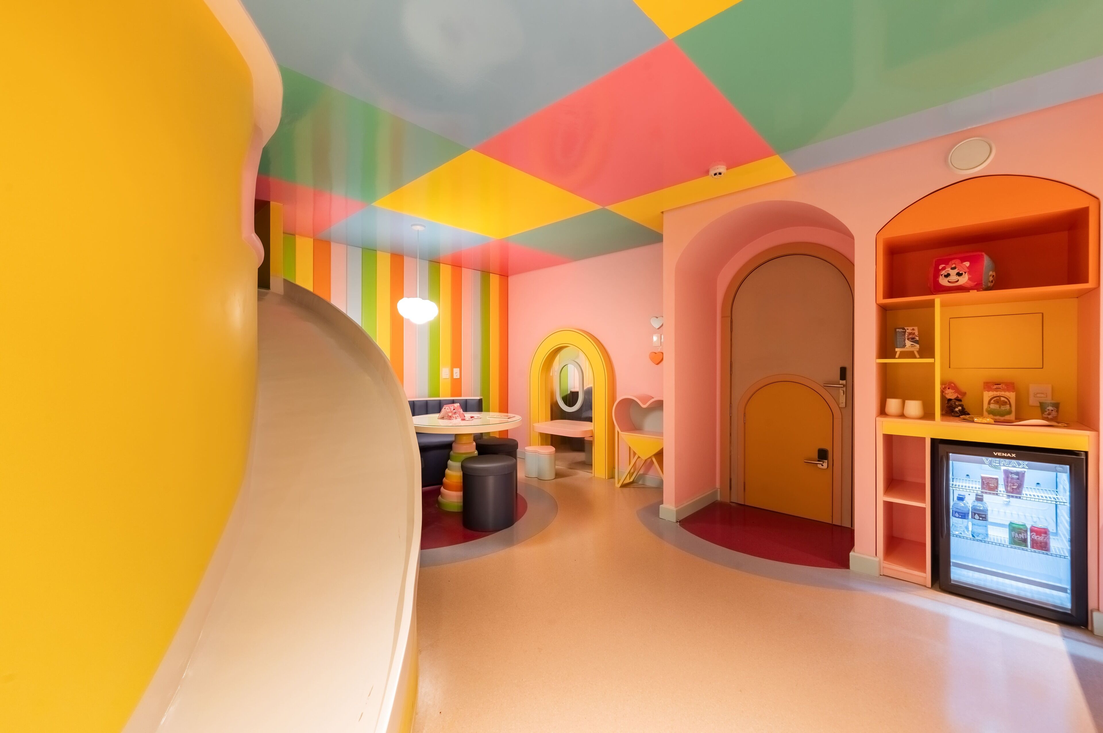 Um espaço de brincadeiras infantil colorido, com um escorrega, uma mesa redonda e uma geladeira.