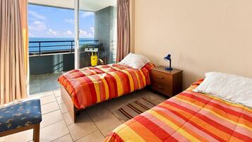 Quarto de hotel com duas camas, uma mesa de cabeceira, uma cadeira e vista para o mar.