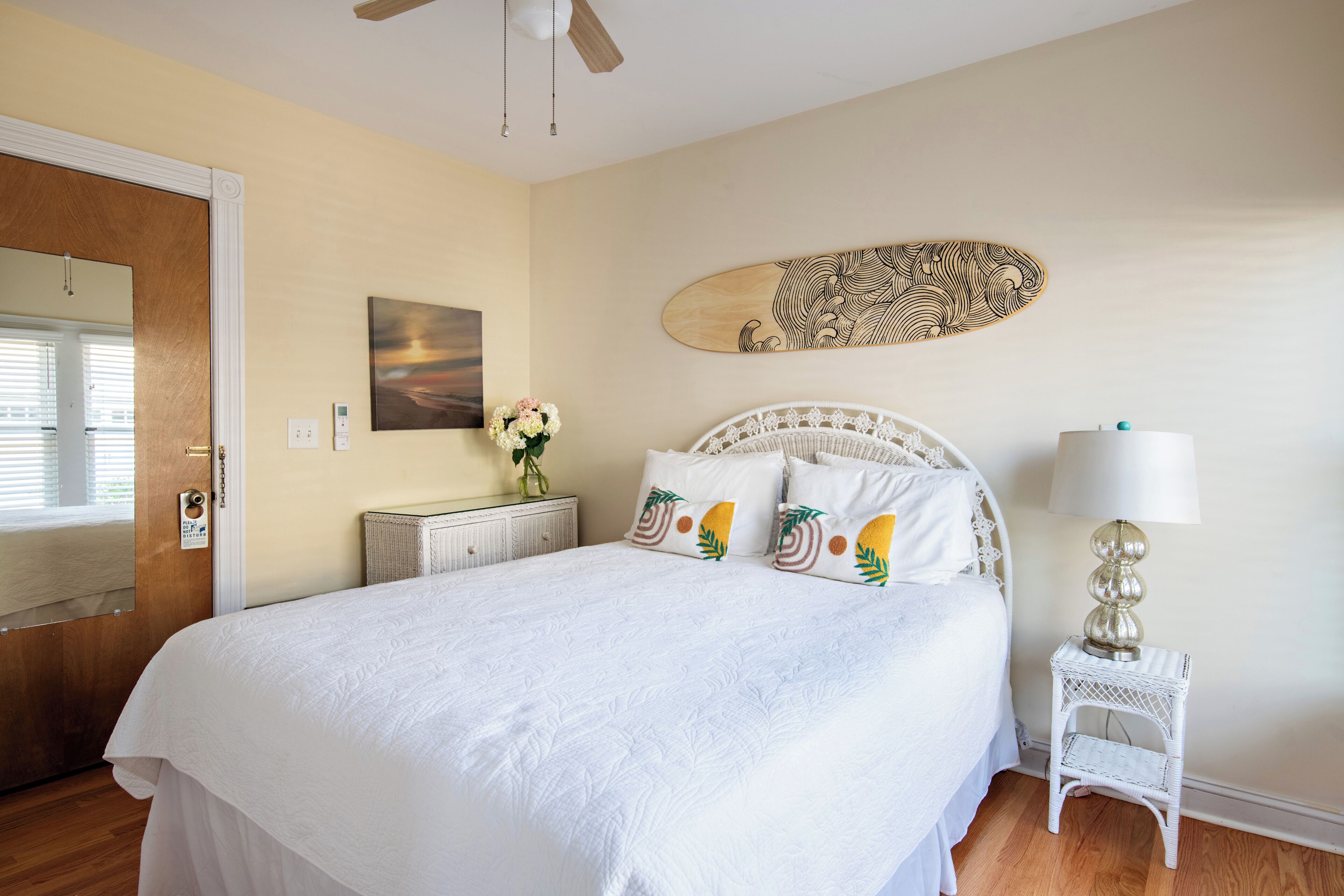Quarto com cama, mesa de cabeceira com abajur, prancha de surf na parede e porta com espelho.