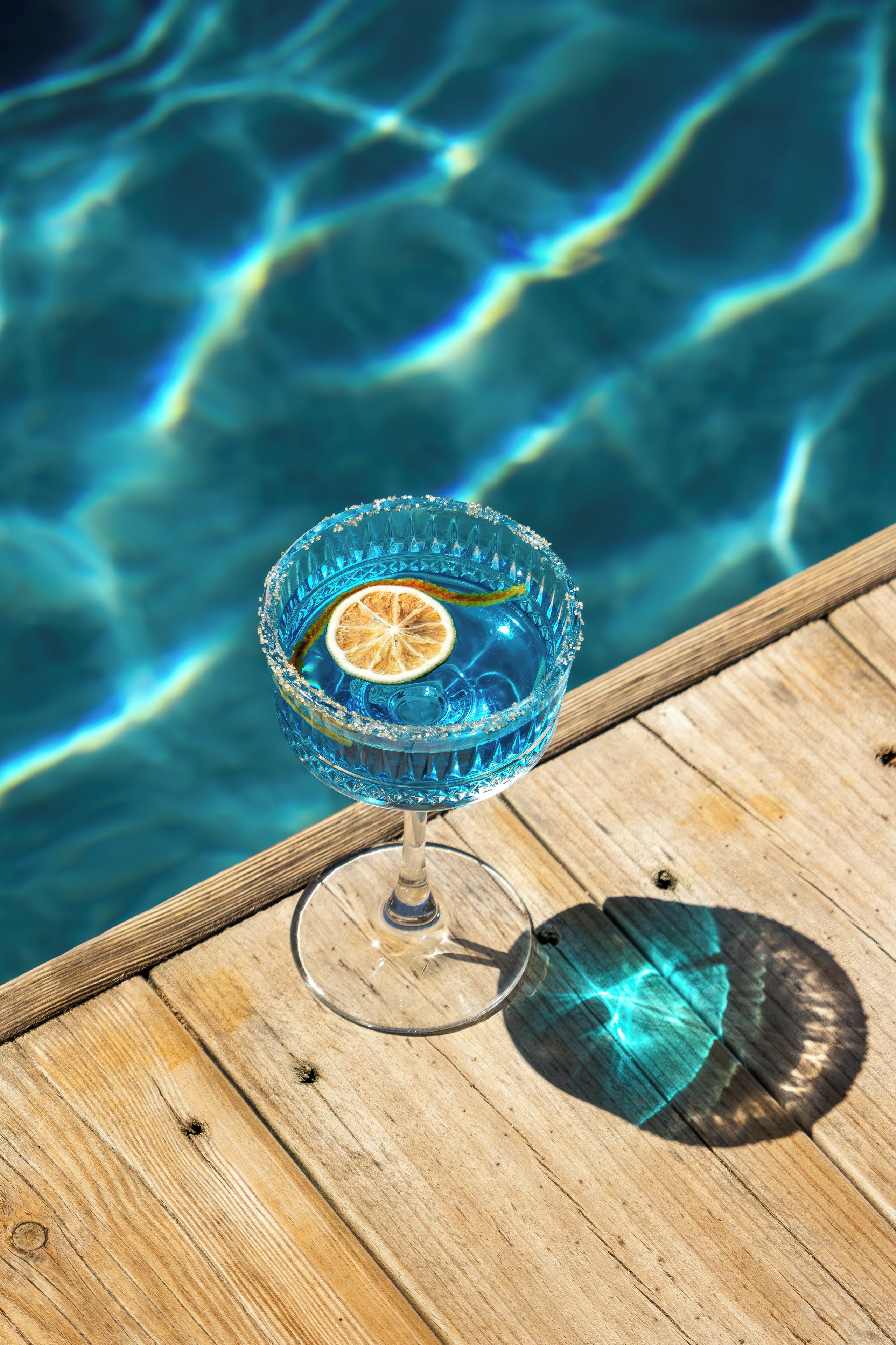 Um copo com base texturizada, uma fatia de fruta cítrica e um líquido transparente, apoiados num deck de madeira junto a uma piscina.