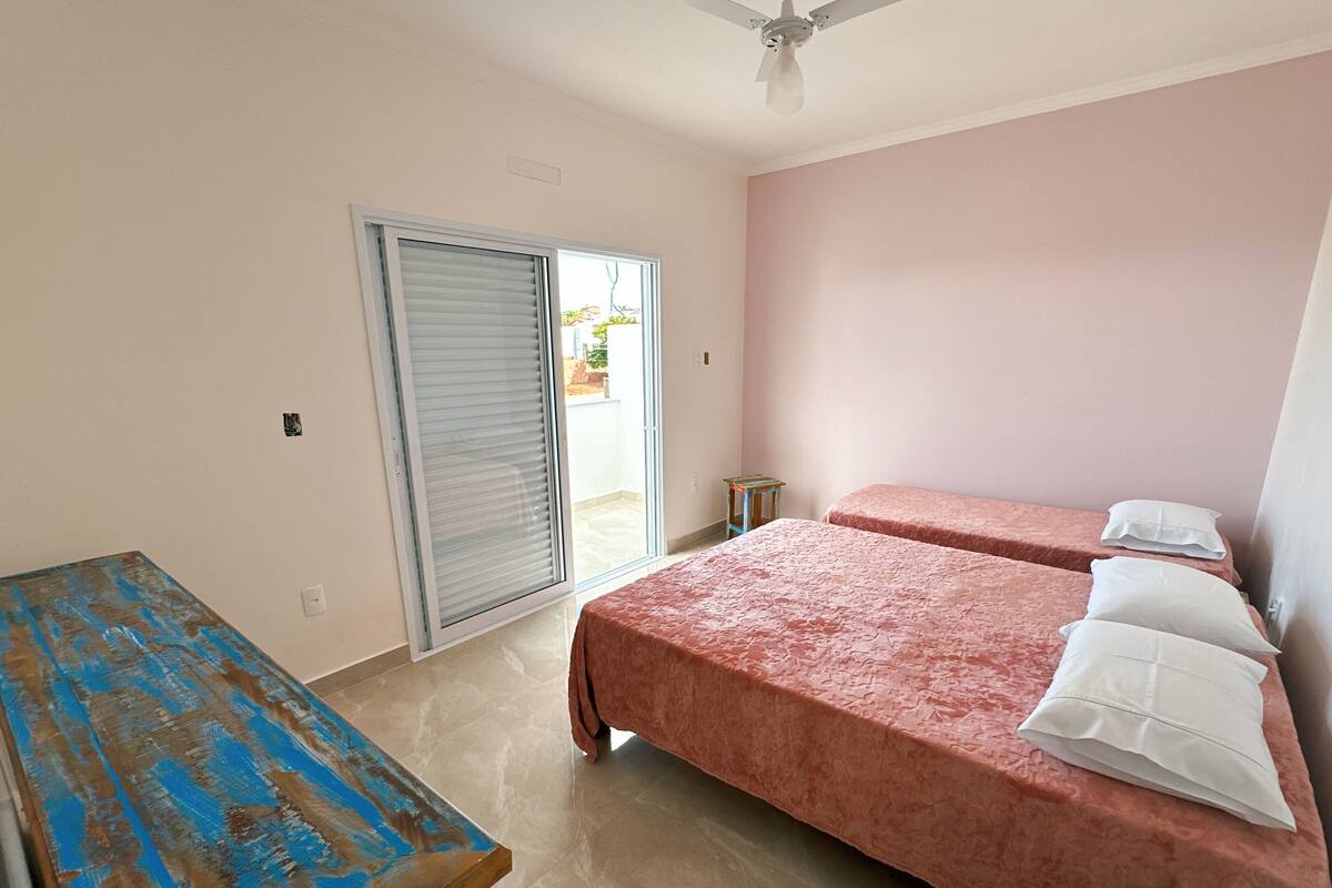 Quarto com duas camas, uma mesa de madeira, um ventilador de teto e uma porta de correr que dá para uma área externa.