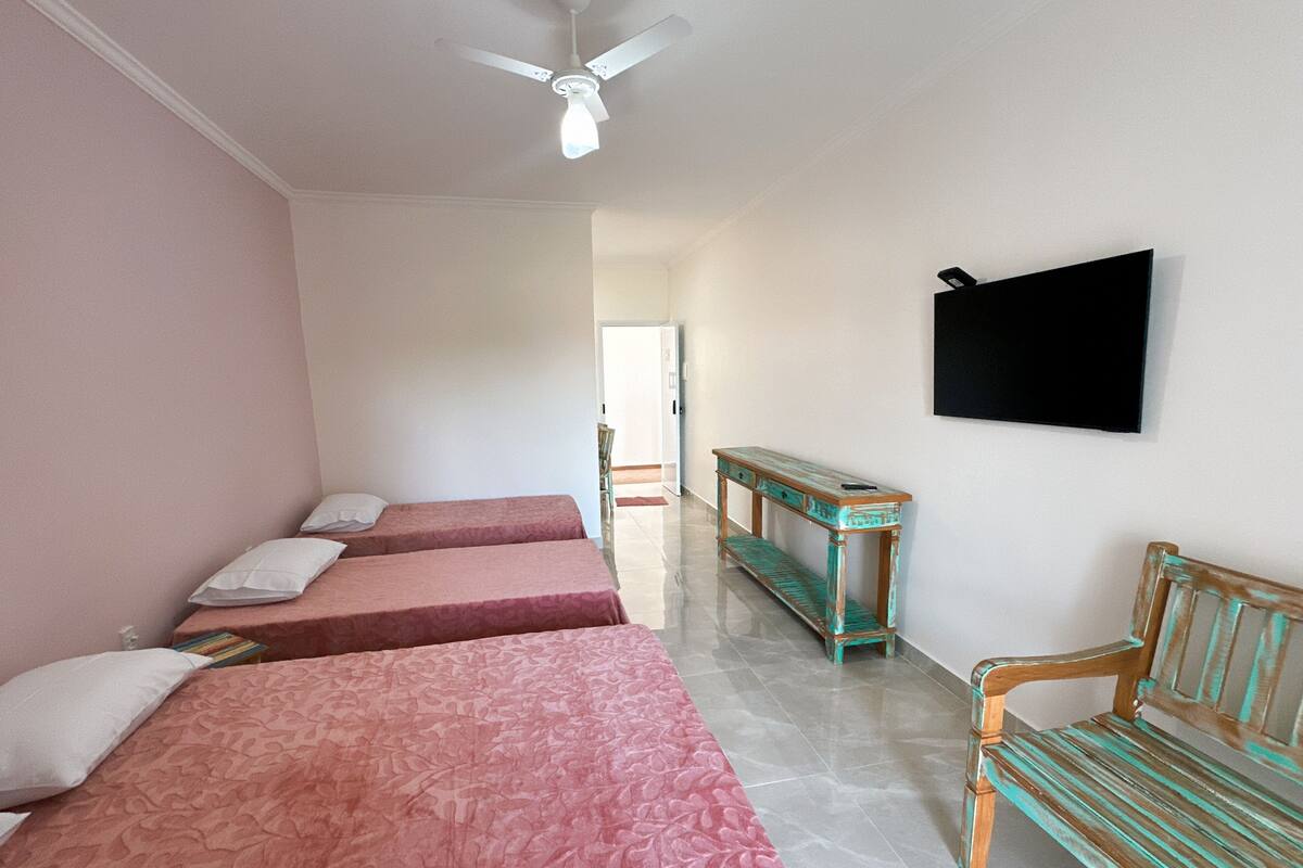 Quarto com três camas, um banco de madeira, uma mesa pequena e um ventilador de teto.