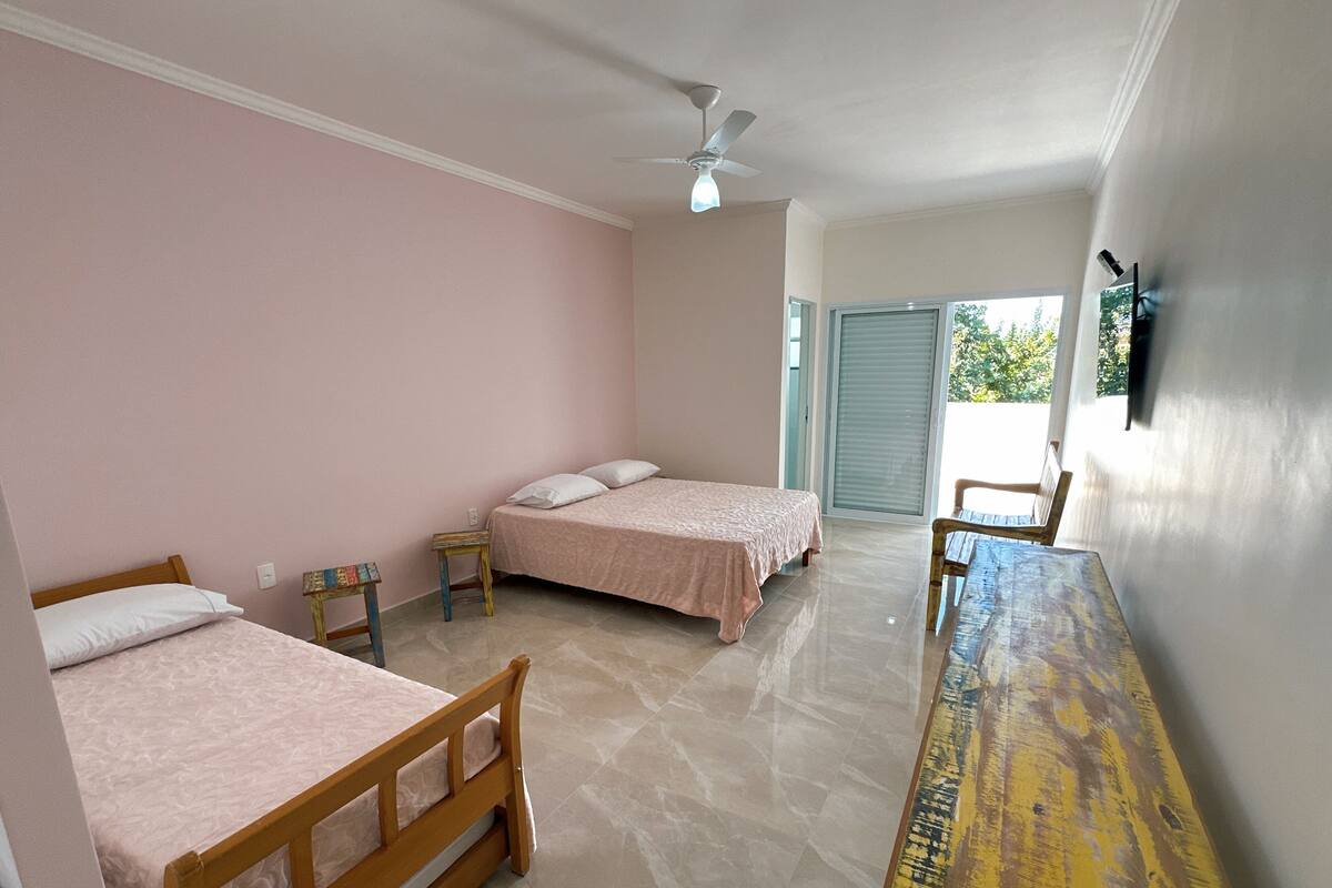 Quarto com duas camas, uma mesa de madeira, um ventilador de teto e vista para o exterior.