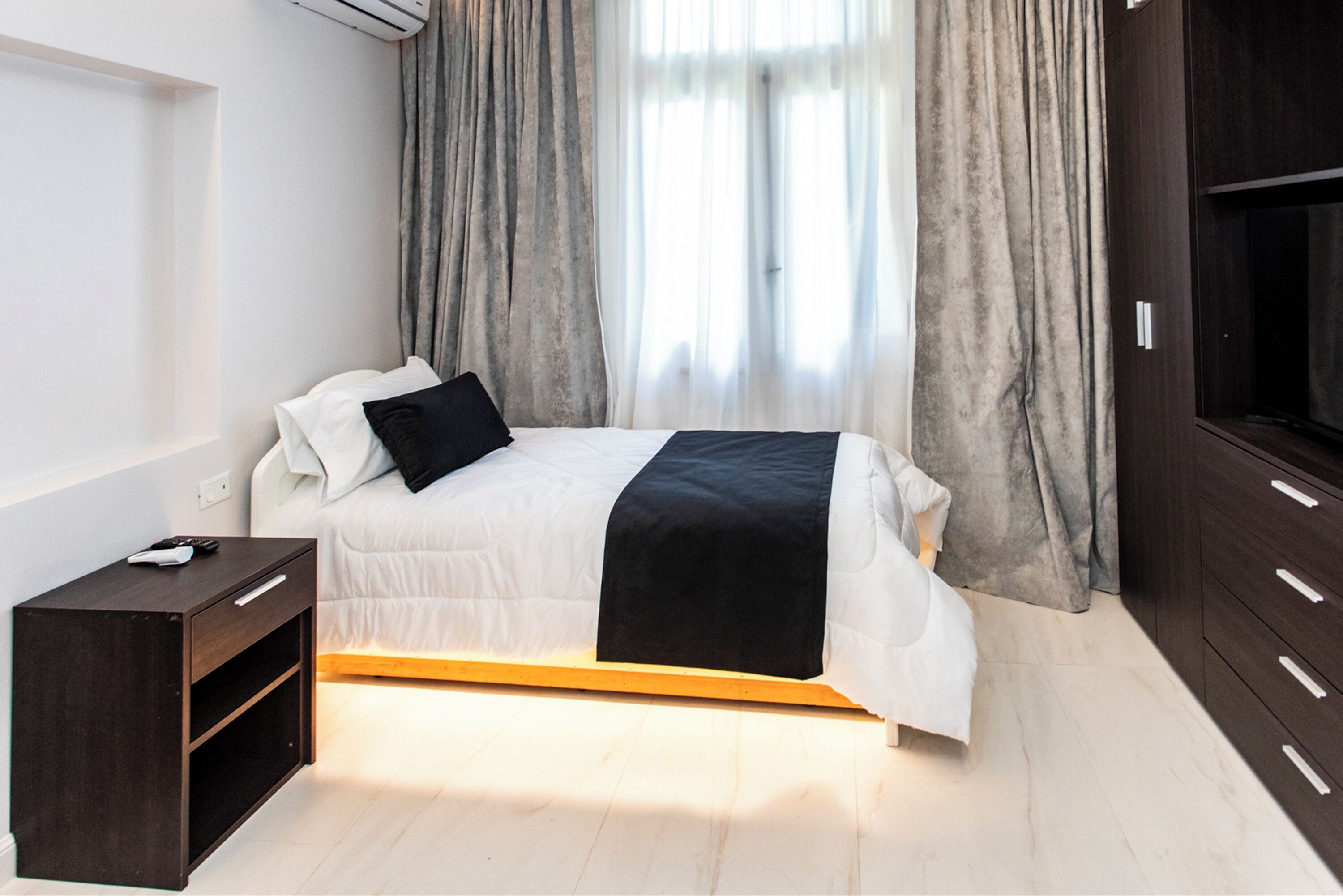 Quarto moderno com cama, mesa de cabeceira, televisão e janela com cortinas.