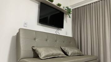 Uma sala de estar moderna com um sofá cinzento, duas almofadas, uma televisão de tela plana na parede e uma estante com objetos decorativos e plantas.