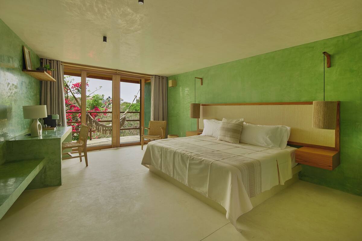 Quarto com uma cama grande, uma escrivaninha com abajur, uma cadeira e vista para uma varanda com plantas.