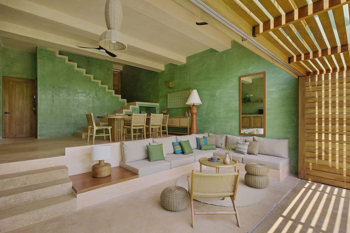 Ampla sala de estar com parede verde, escada, conjunto de sofás e área de jantar com móveis de vime.