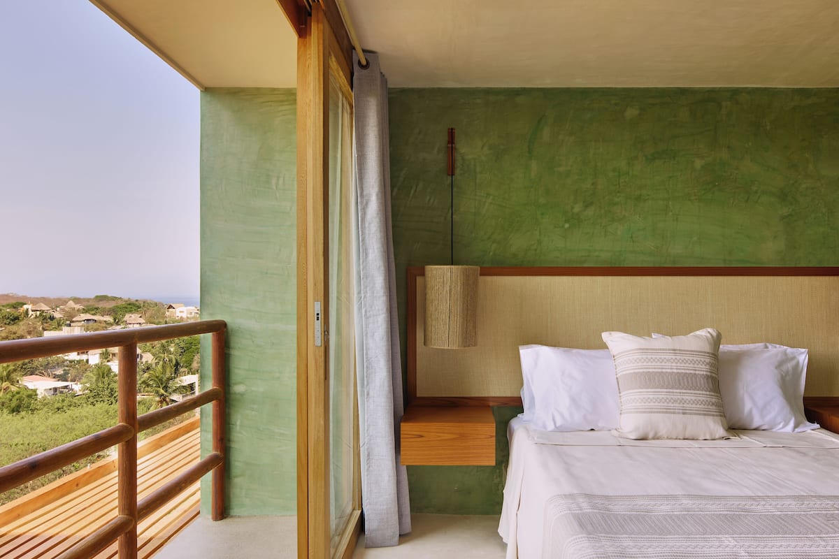 Varanda com guarda de madeira, cama com roupa de cama branca e almofada estampada, luminária pendente e vista para uma paisagem verde e edifícios.