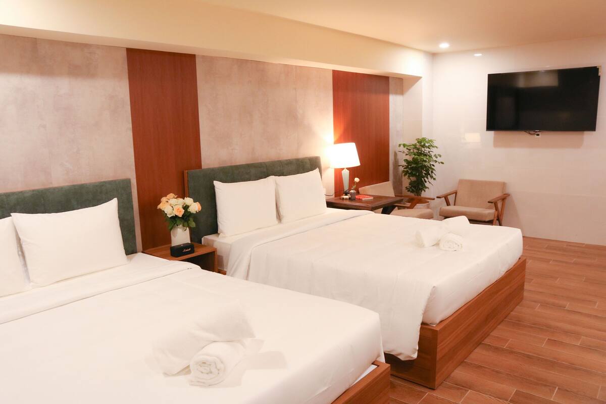 Quarto de hotel com duas camas, televisão, uma cadeira e mesa de cabeceira com luminária e flores.