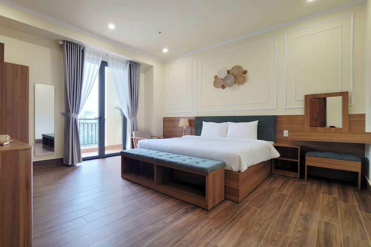Quarto de hotel com piso de madeira, cama com cabeceira verde, escrivaninha, um banco e uma janela grande com cortinas.