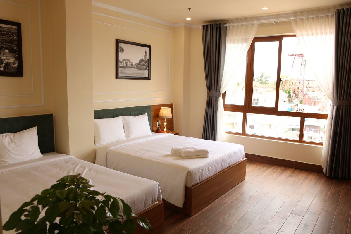 Quarto de hotel com duas camas, uma janela grande com cortinas, um quadro na parede e uma planta no canto.