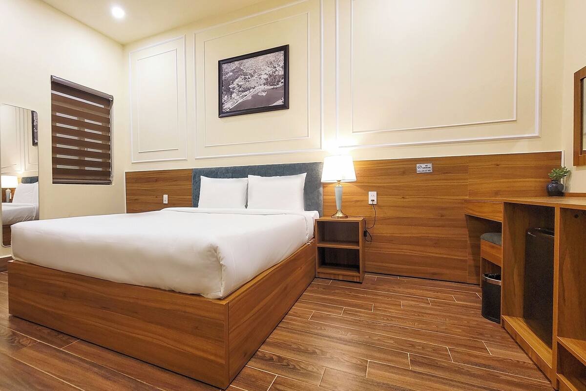 Quarto de hotel com uma cama de madeira, mesa de cabeceira com abajur, pequena escrivaninha e um quadro pendurado na parede.