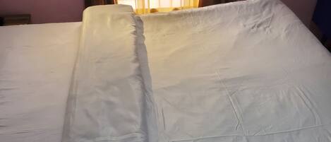 Uma cama com lençol branco em um quarto com parede roxa e uma janela com cortinas.