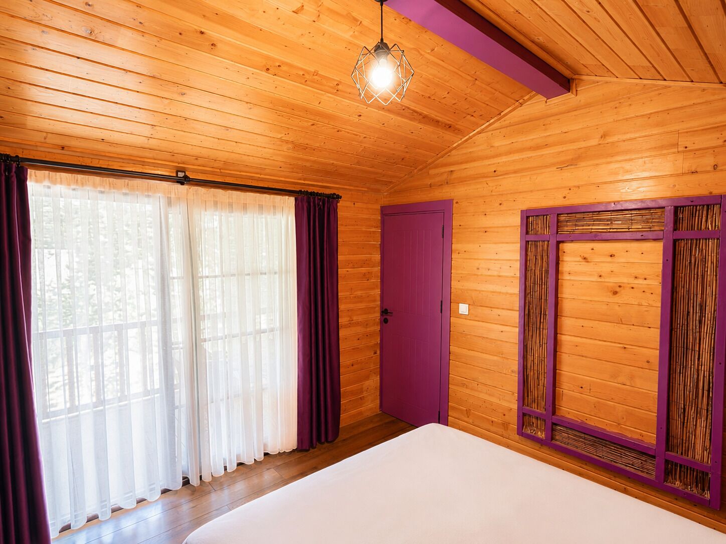 Um quarto de madeira com uma cama, uma porta roxa e uma janela com cortinas leves.