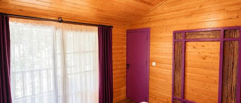 Um quarto de madeira com uma cama, uma porta roxa e uma janela com cortinas leves.