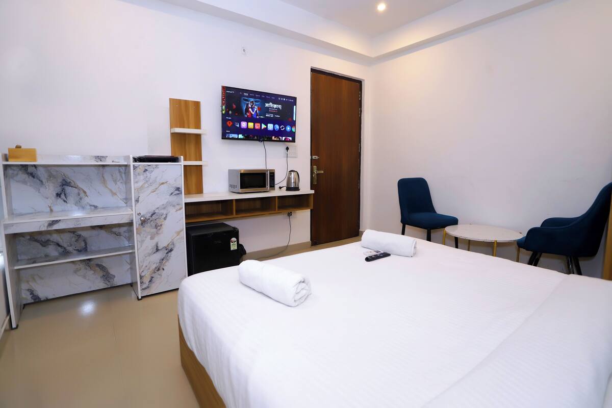 Quarto de hotel com cama, televisão na parede, micro-ondas, mesa pequena e duas cadeiras.