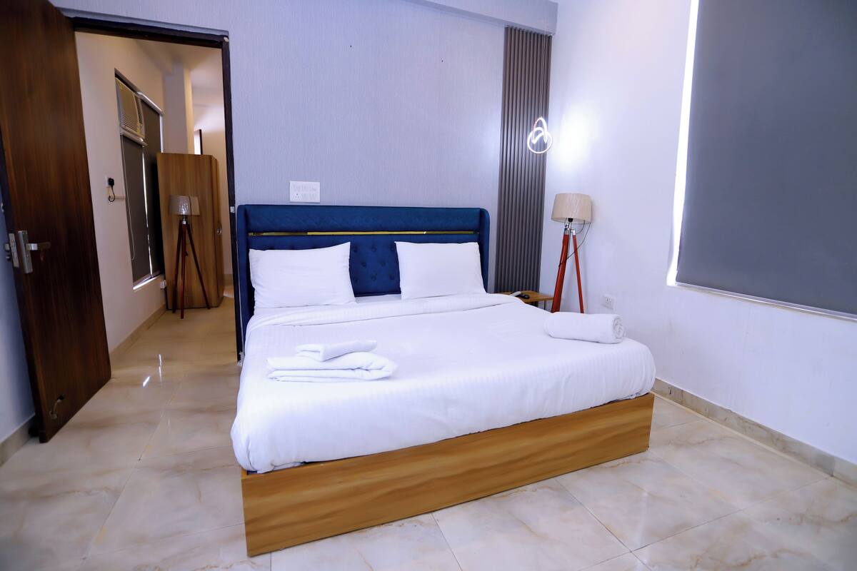 Cama bem arrumada com lençóis brancos e cabeceira azul, luminária de cabeceira e vista para o corredor.