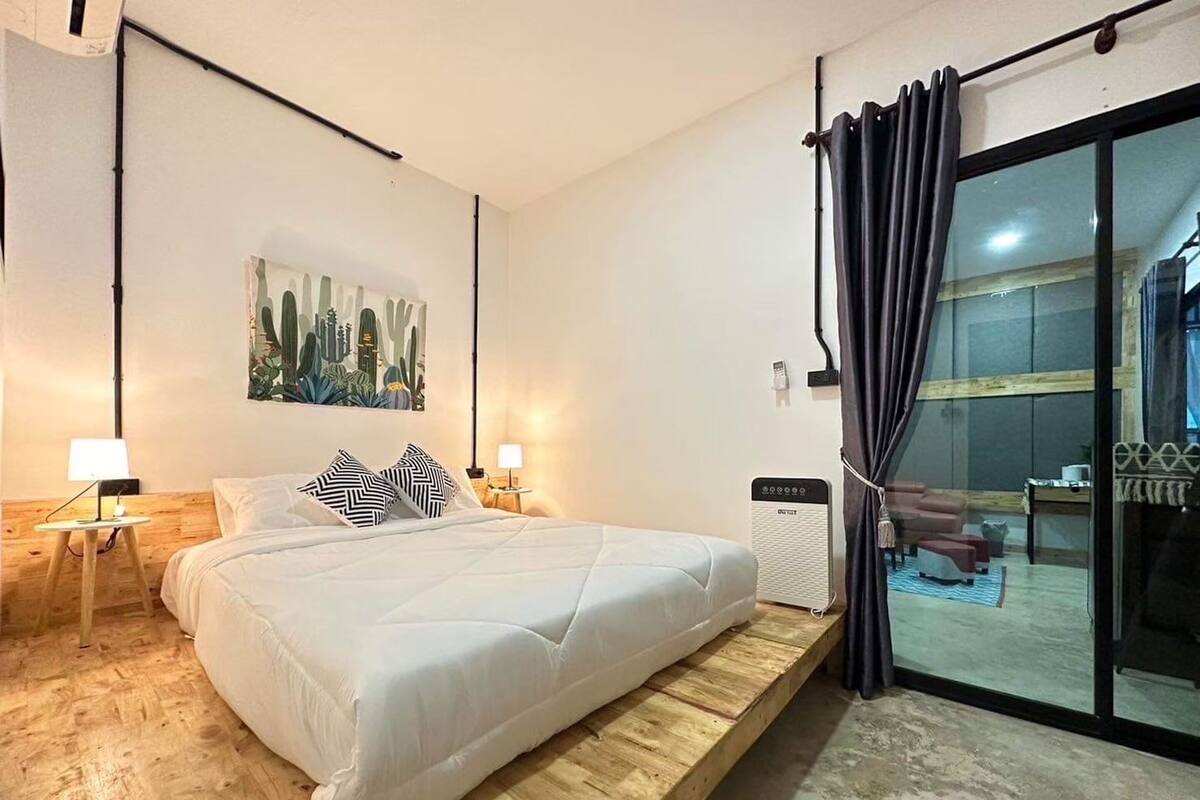 Quarto com cama, mesa de cabeceira, abajur, arte na parede com cactos, aquecedor e porta de vidro deslizante para uma varanda.