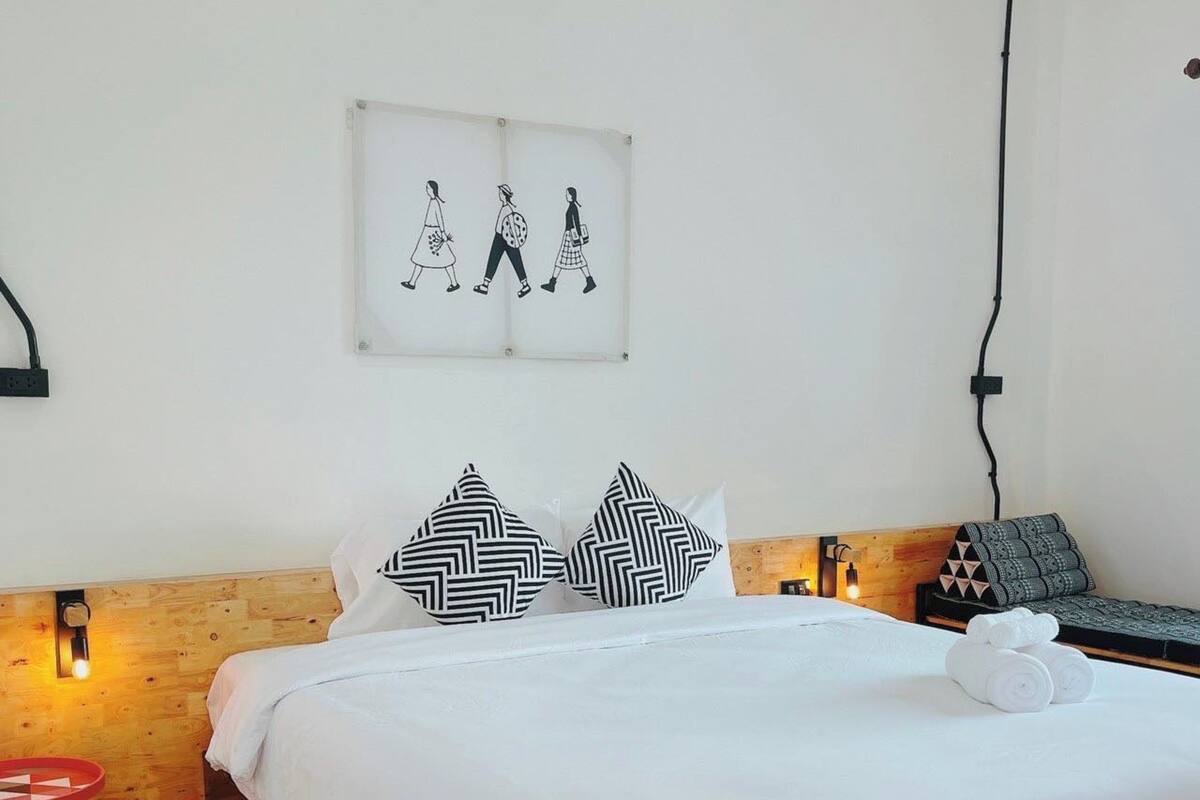 Quarto de hotel com uma cama grande, duas almofadas padronizadas em preto e branco, uma obra de arte na parede retratando três pessoas caminhando, uma cabeceira de madeira e um banquinho vermelho pequeno.