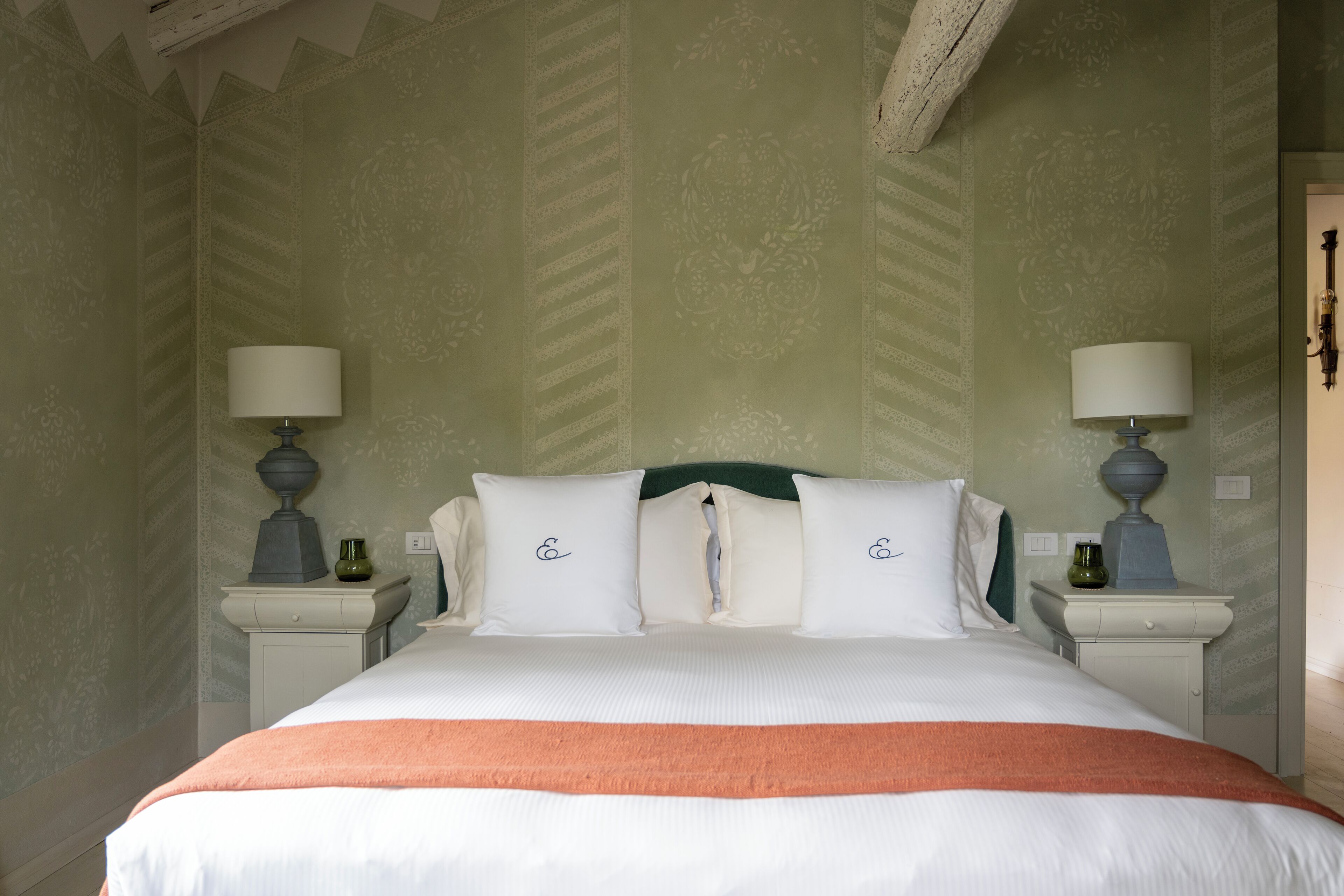 Cama bem arrumada com roupa de cama branca e laranja, duas mesas de cabeceira com abajures e papel de parede estampado.