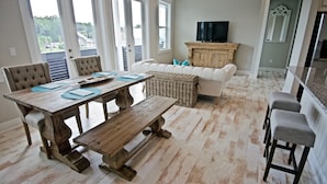 Uma sala de estar ampla com uma mesa de jantar de madeira, assentos acolchoados, uma televisão e uma área de cozinha.