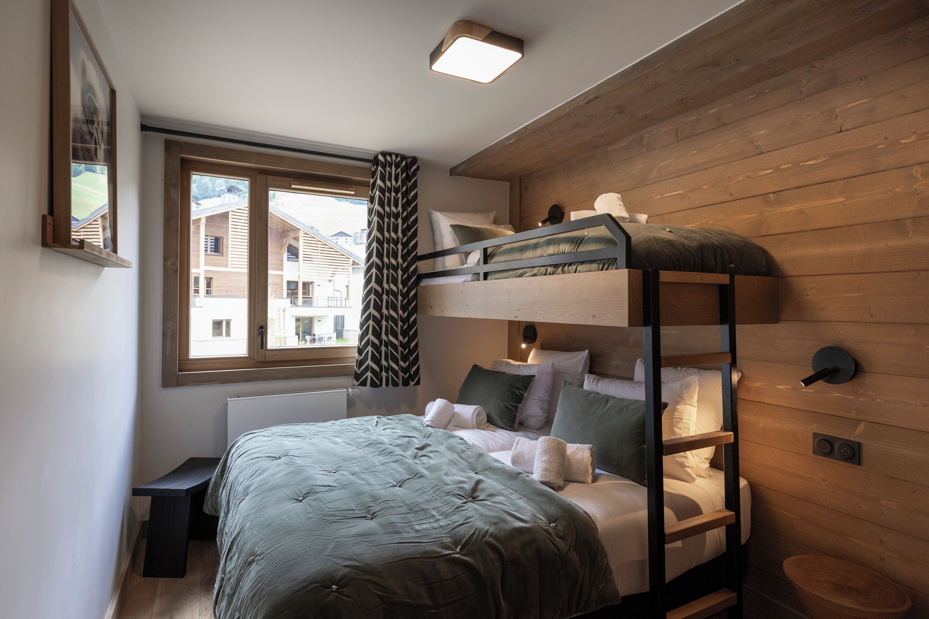 Quarto com beliche e vista para edifícios, janela com cortinas e mesa de cabeceira.