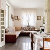Uma sala de jantar com mesa posta para quatro pessoas, um sofá com um colcha colorida e uma janela com cortinas leves.