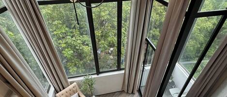 Apartamento alto com grande janela oferecendo vista da cidade, área de estar aconchegante com sofá e uma pequena mesa, e escada que leva a um andar superior.