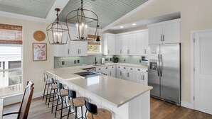 Uma cozinha moderna com armários brancos, uma ilha grande com banquetas e uma geladeira de aço inoxidável, além de iluminação pendente.