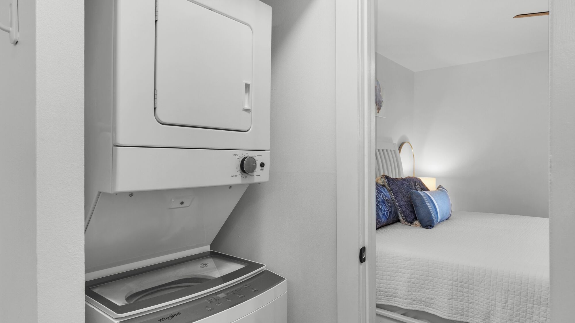Uma área de lavanderia com máquina de lavar e secadora frontais, uma cama com colcha branca e uma mesa de cabeceira com luminária e almofadas.