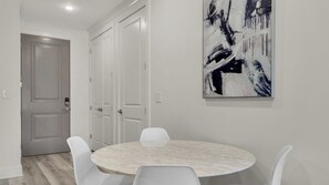 Uma área de refeições moderna com uma mesa redonda de mármore e cadeiras brancas, um grande quadro abstrato na parede e um conjunto de portas brancas ao fundo.