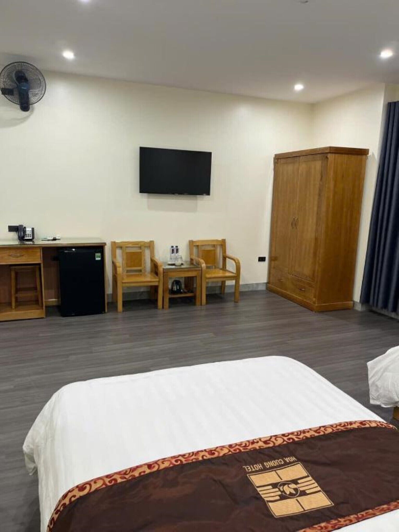 Quarto de hotel com uma cama, uma escrivaninha com computador, duas cadeiras, uma televisão e um guarda-roupa.