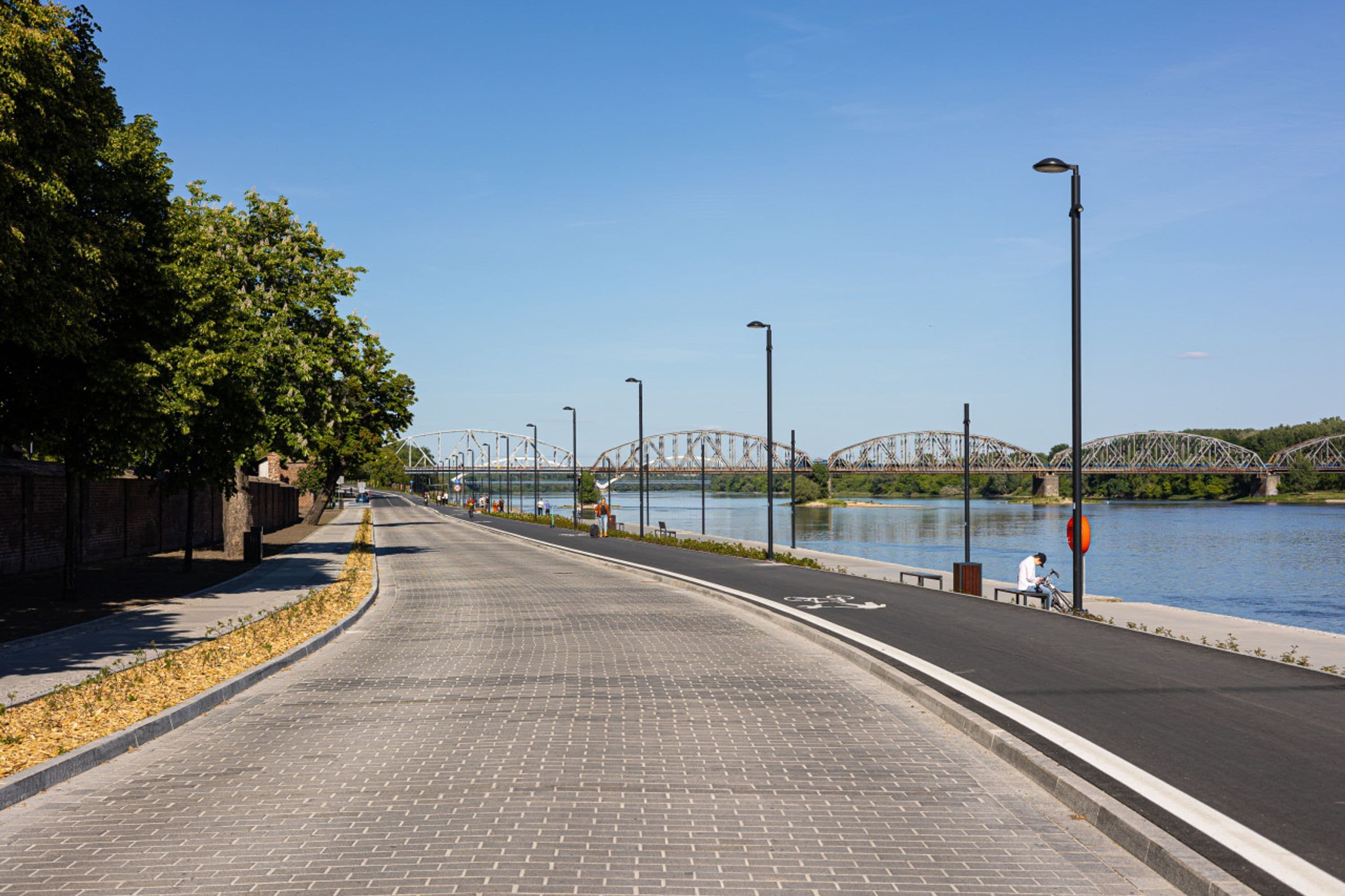 Uma passarela pavimentada ao longo de um rio, com uma ponte ao fundo.