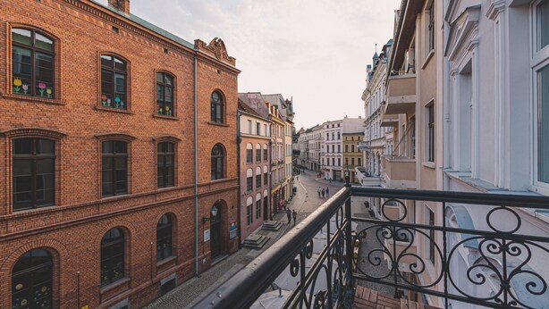 Uma rua estreita com edifícios históricos, um prédio de tijolos com janelas em arco e uma varanda com grades ornamentadas.