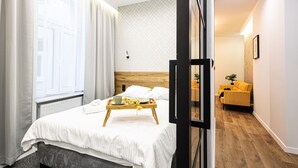 Quarto de hotel moderno com uma cama grande, cabeceira de madeira, mesa de cabeceira com um tabuleiro e vista para um corredor com um sofá amarelo e uma planta.