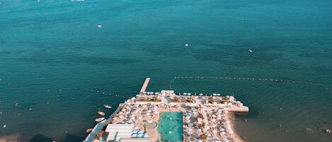 Vista aérea de um resort costeiro com piscina, guarda-sóis de praia e um píer que se estende até o mar.